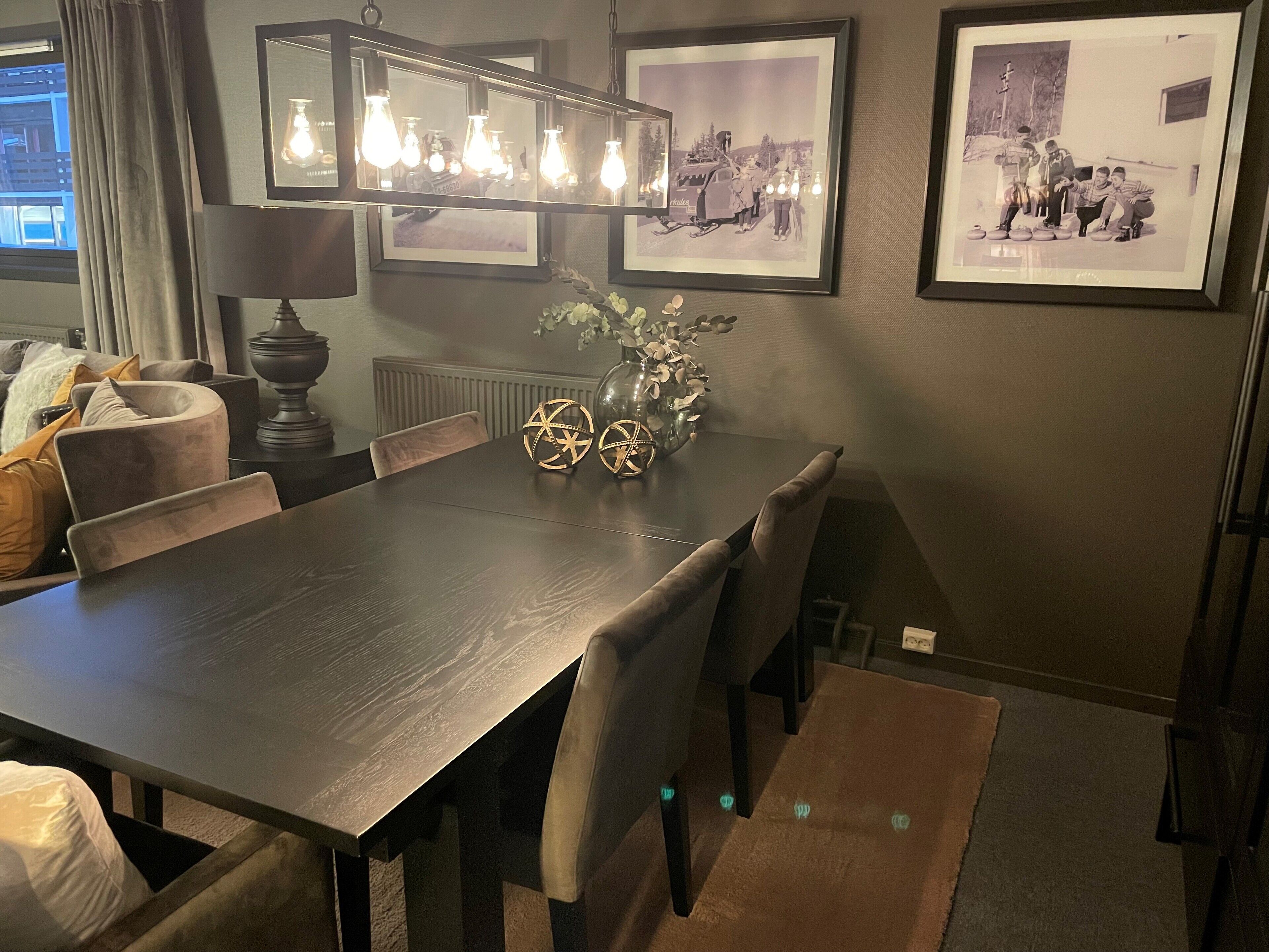 Uma sala de jantar com uma mesa de madeira escura, cadeiras acolchoadas, um vaso com flores e fotografias emolduradas na parede.
