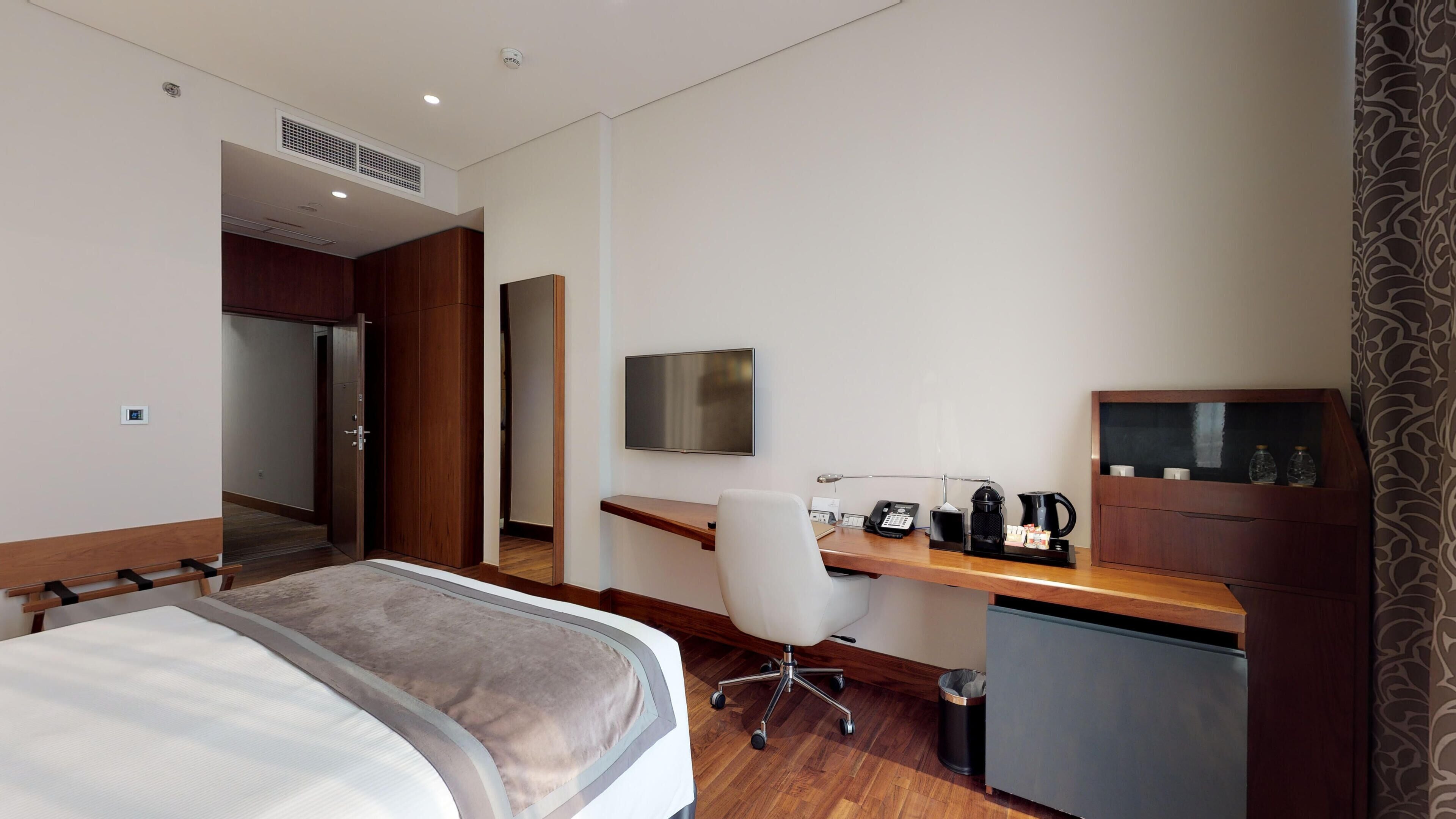 Quarto de hotel com uma cama, uma escrivaninha com cadeira, televisão, máquina de café e um pequeno frigorífico.