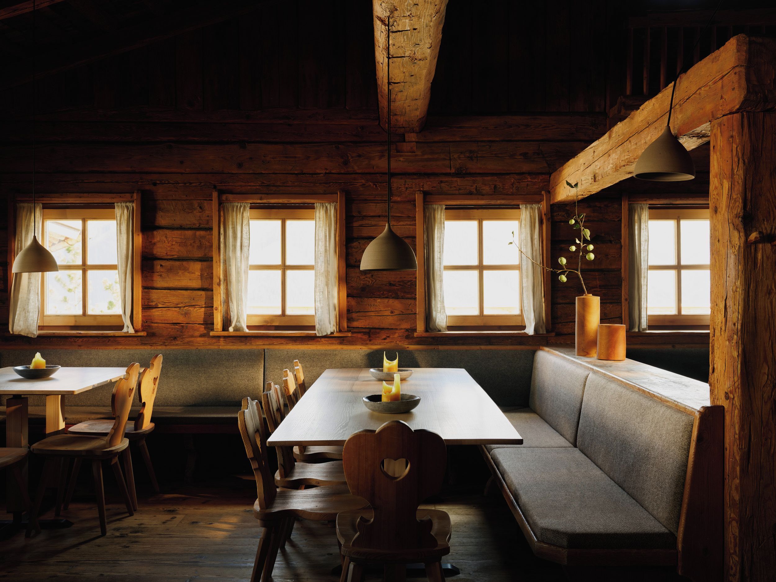 Interior rústico de uma cabana de madeira, com uma mesa de jantar, cadeiras, um banco e duas janelas com cortinas.