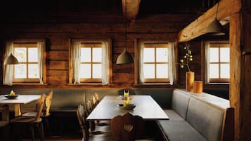 Interior rústico de uma cabana de madeira, com uma mesa de jantar, cadeiras, um banco e duas janelas com cortinas.