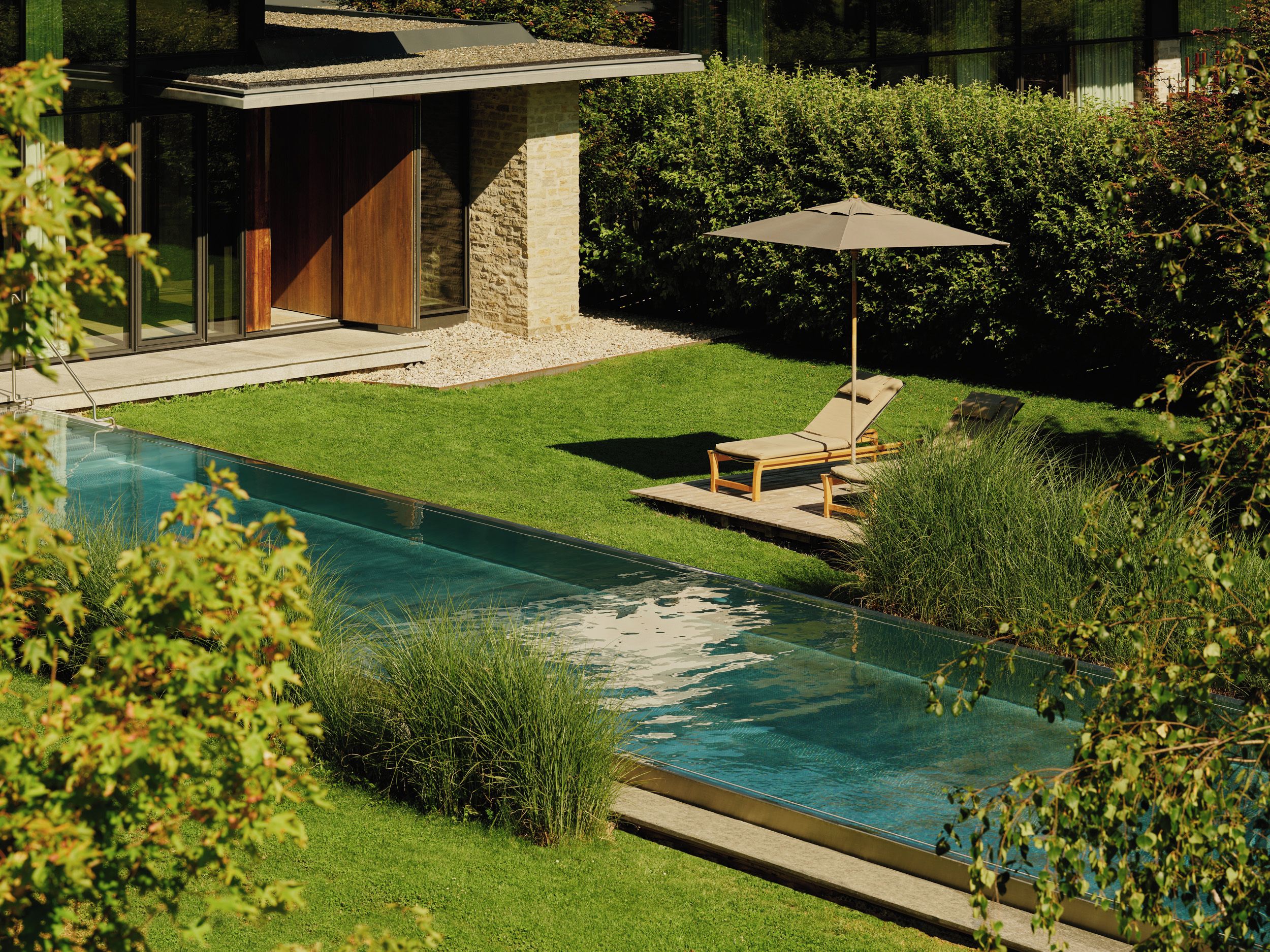 Piscina com espreguiçadeira e guarda-sol, cercada por gramado e arbustos bem cuidados.