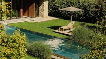 Piscina com espreguiçadeira e guarda-sol, cercada por gramado e arbustos bem cuidados.