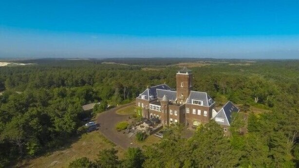 Um edifício grande e histórico com uma torre imponente, rodeado por uma floresta densa.