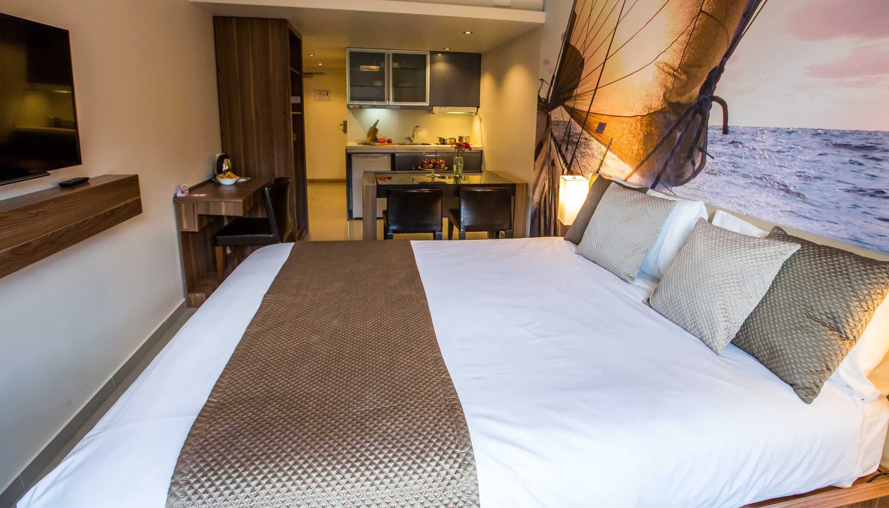 Quarto de hotel com uma cama grande, uma escrivaninha e uma kitchenette ao fundo.