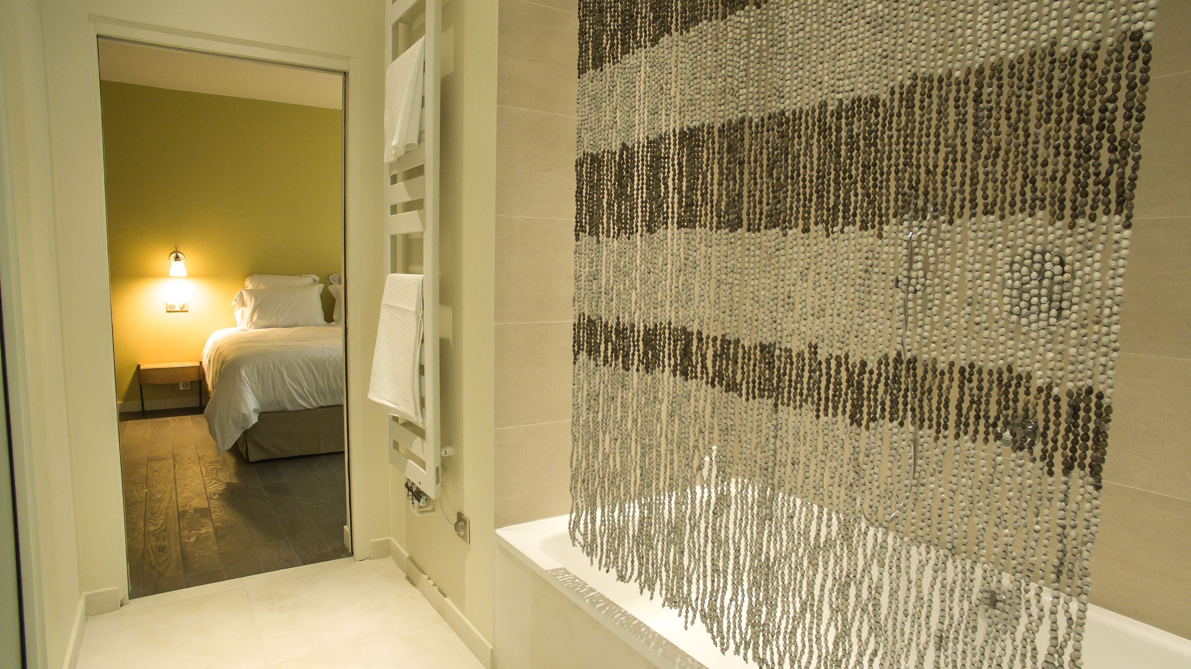 Um banheiro com cortina de chuveiro feita de contas, uma cama com lençóis brancos e uma mesa de cabeceira com abajur.