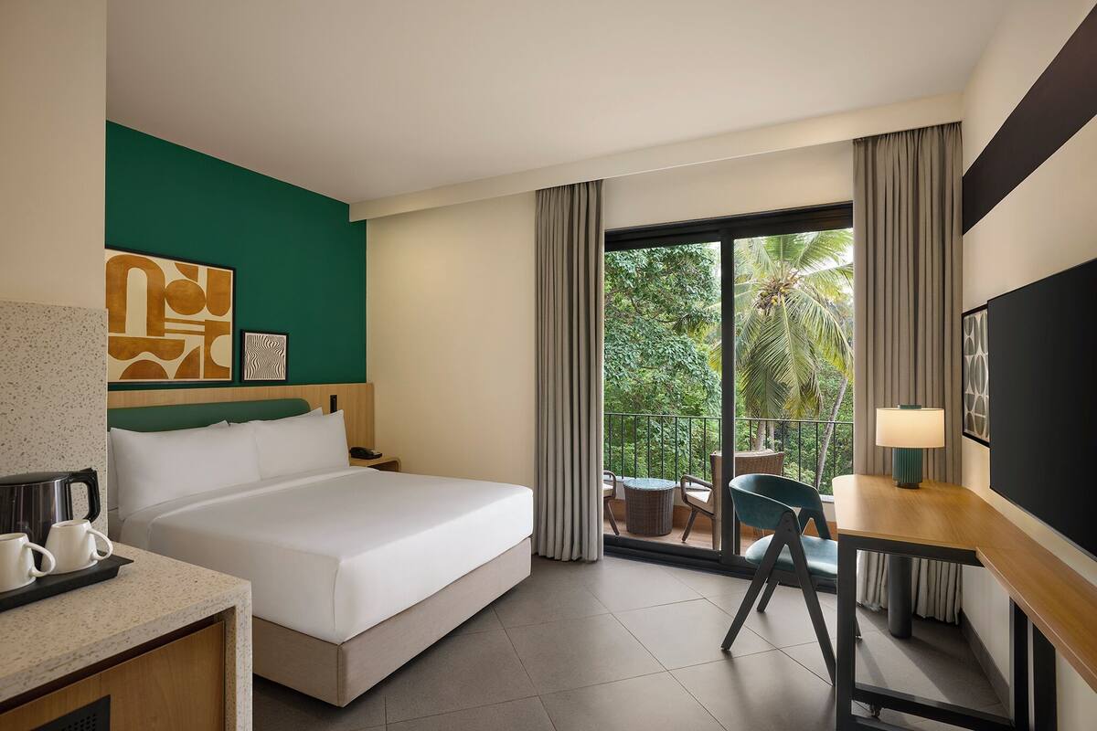 Quarto de hotel com cama, escrivaninha, cadeira, televisão, máquina de café e varanda com vista para o verde.