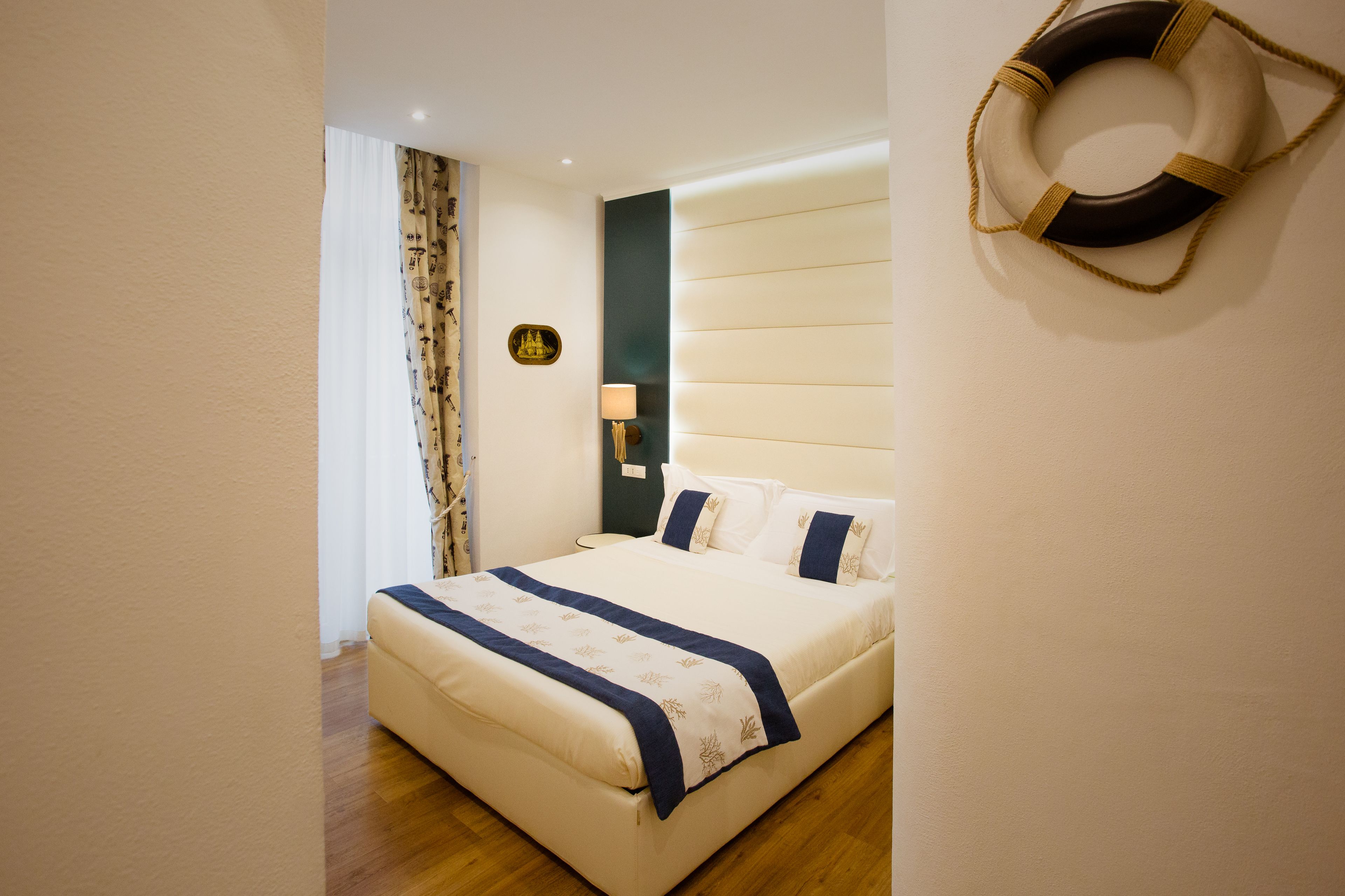 Cama bem arrumada com colcha branca e azul-marinho, abajur e um círculo salva-vidas pendurado na parede.