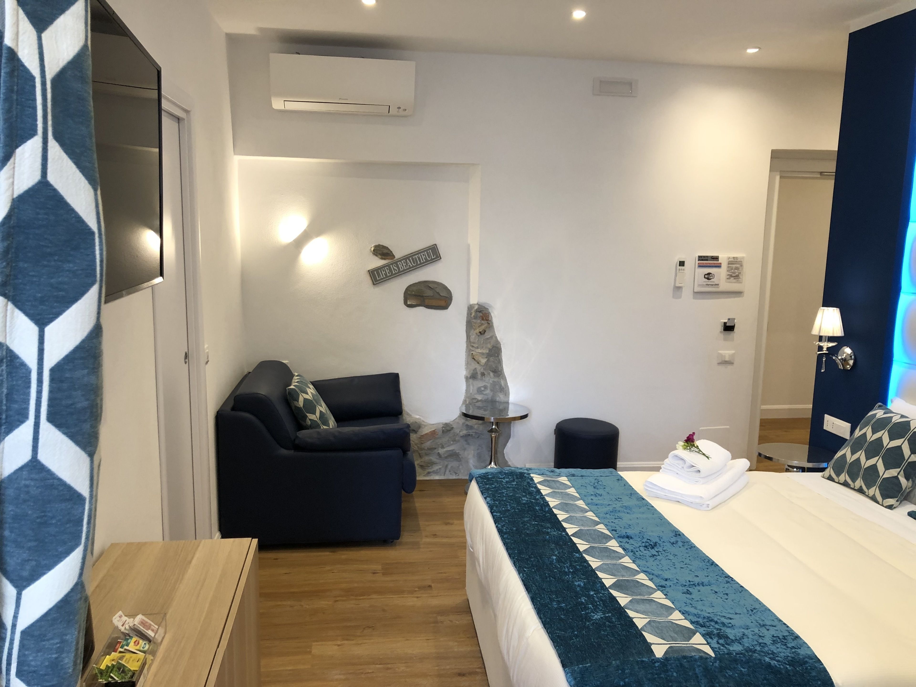 Quarto de hotel com cama, sofá, pequena mesa, unidade de ar condicionado, parede decorativa de pedra e luminária fixada na parede.
