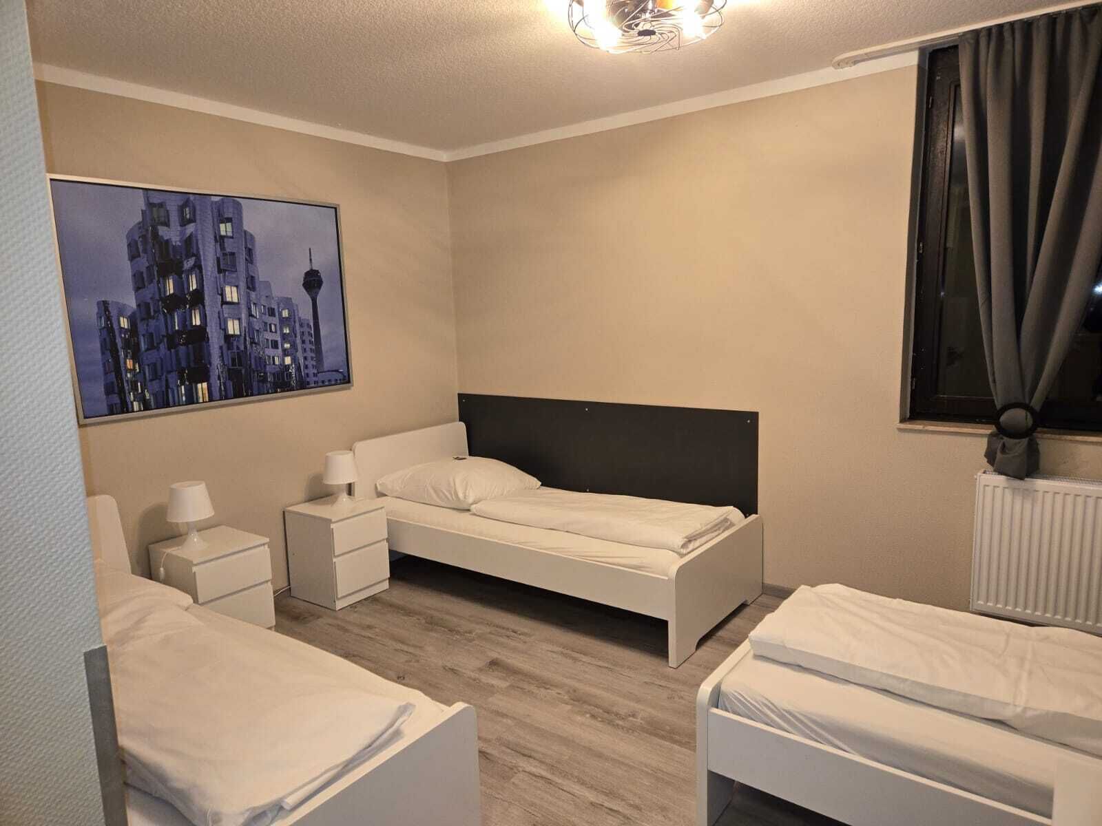 Quarto de hotel com uma cama, uma mesa de cabeceira, uma luminária, uma janela com cortinas e uma pintura de paisagem urbana na parede.