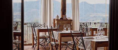 Um restaurante com área externa, mesas com toalhas brancas e copos de vinho, e vista para uma paisagem montanhosa.