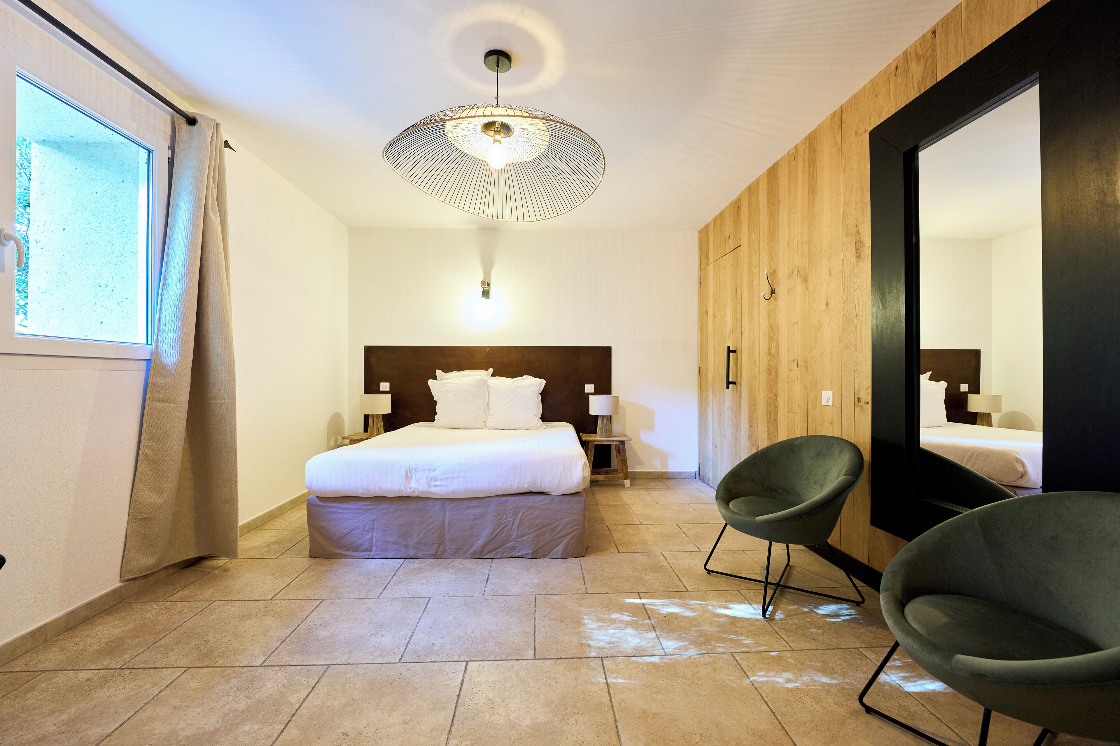 Quarto de hotel com cama, duas luminárias de cabeceira, uma cadeira, um espelho grande e um lustre.