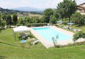 Uma piscina retangular com um toldo branco e espreguiçadeiras ao redor, situada em uma área gramada com árvores e vista para colinas ao longe.