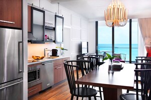 Uma cozinha moderna com eletrodomésticos de aço inoxidável, uma área de refeições com mesa de madeira e cadeiras pretas, e uma grande janela com vista para o oceano.