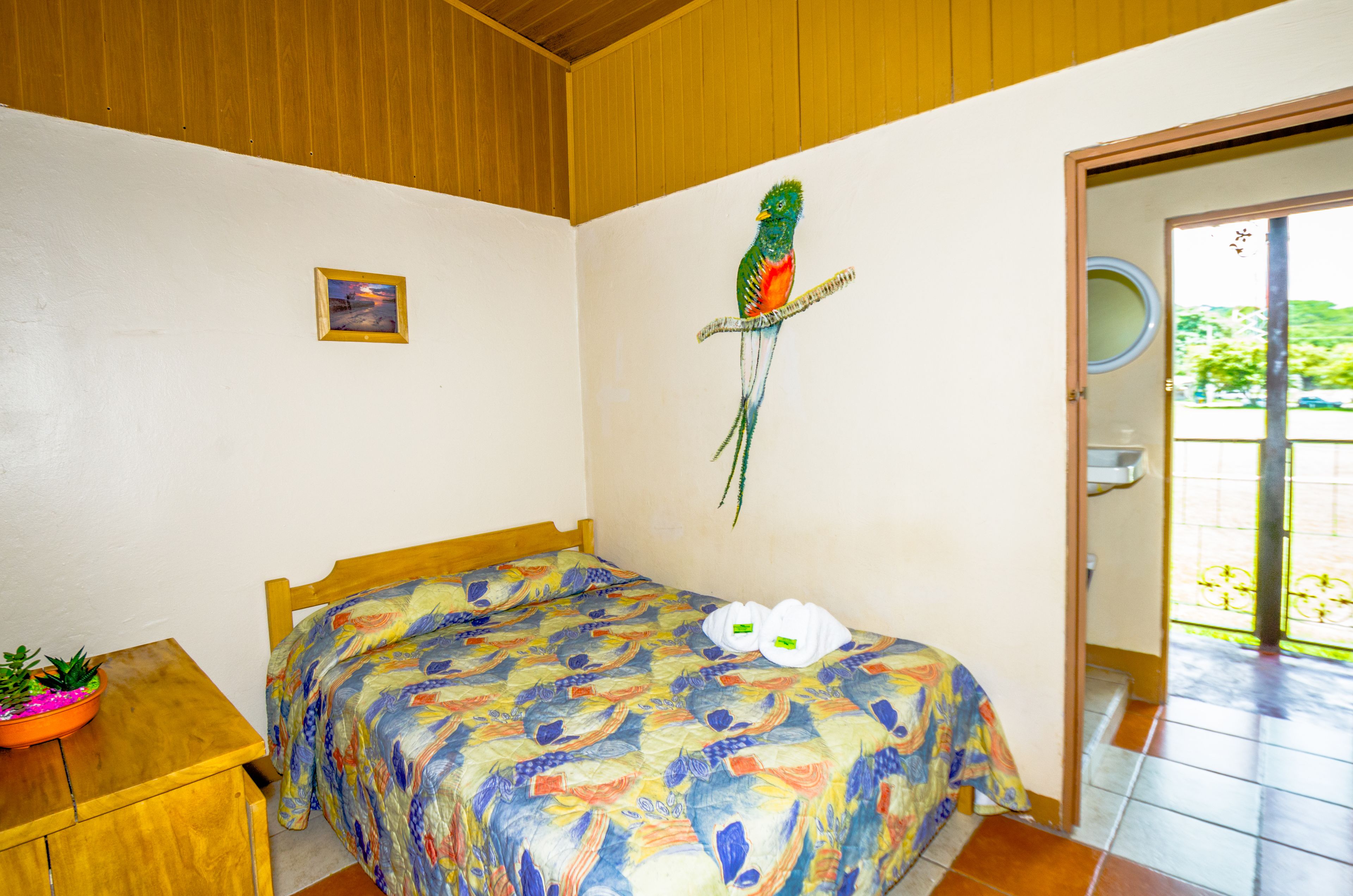 Quarto com cama, cabeceira de madeira, mesinha com planta, pintura de um pássaro e porta que dá para uma varanda.