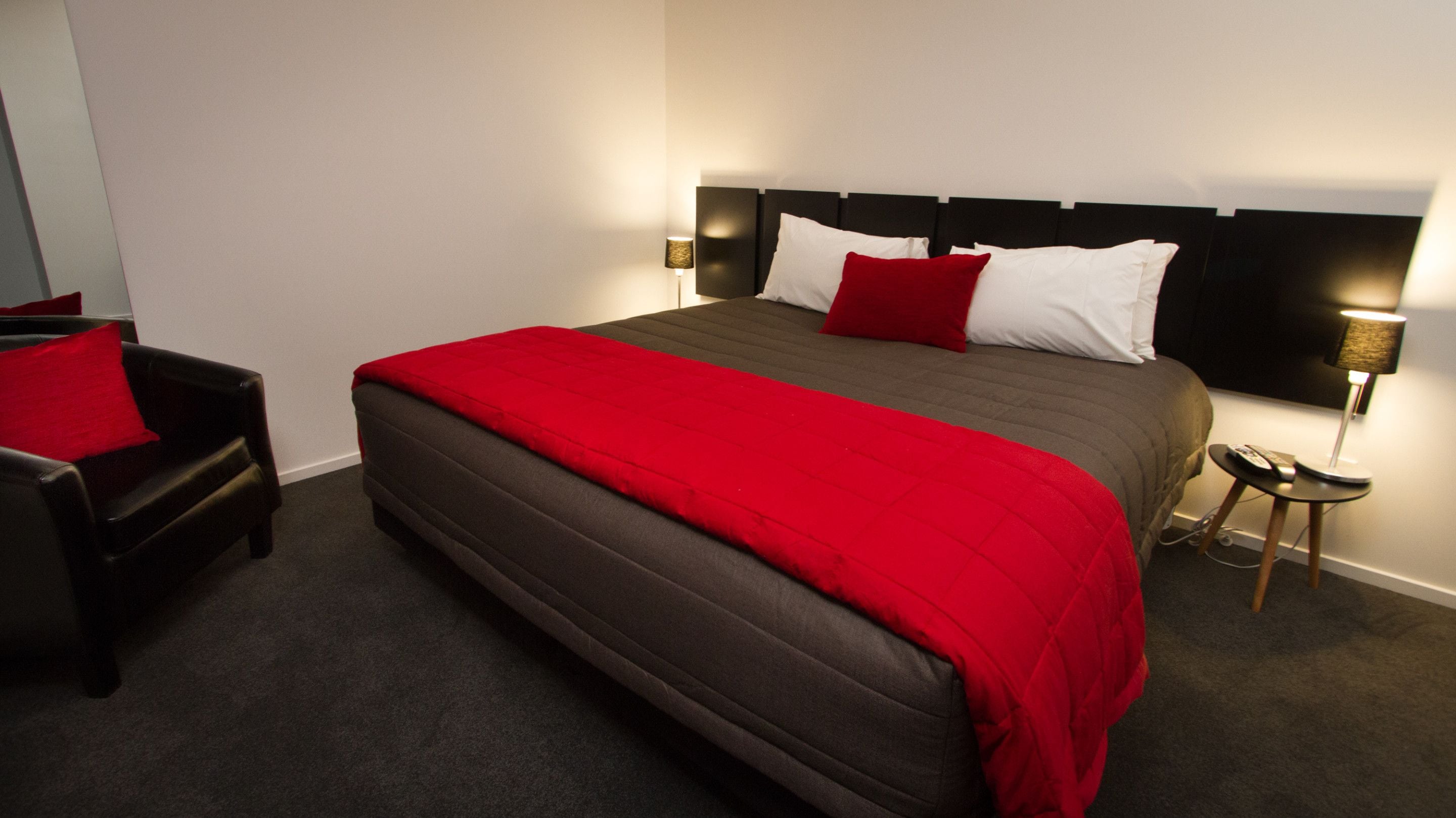 Quarto de hotel com uma cama grande, um cobertor vermelho, cabeceira preta, mesa de cabeceira com abajur e uma poltrona de couro preta.