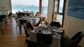 Uma sala de jantar com mesas para quatro, grandes janelas com vista para o mar e piso de madeira.