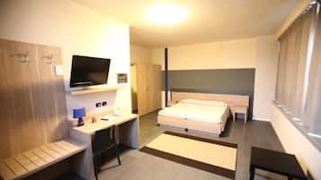 Quarto de hotel com cama, escrivaninha, cadeira, televisão e janela com cortinas.