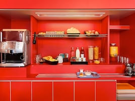 Bancada de café da manhã com tema vermelho, incluindo máquina de café, pão, doces e diversas opções para o café.