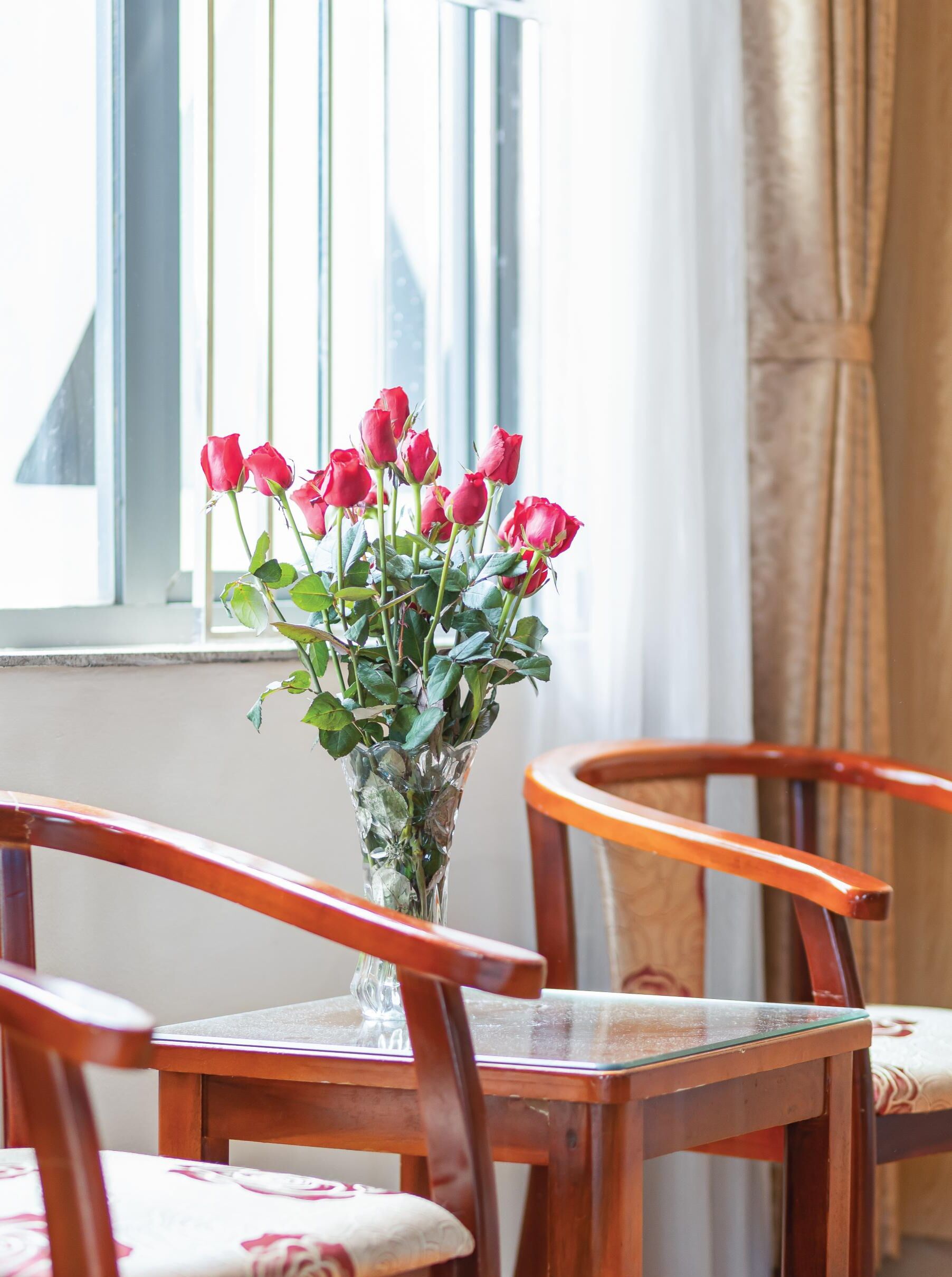 Uma cadeira de madeira com almofada, um vaso transparente com rosas cor-de-rosa, e uma janela com persianas verticais ao fundo.