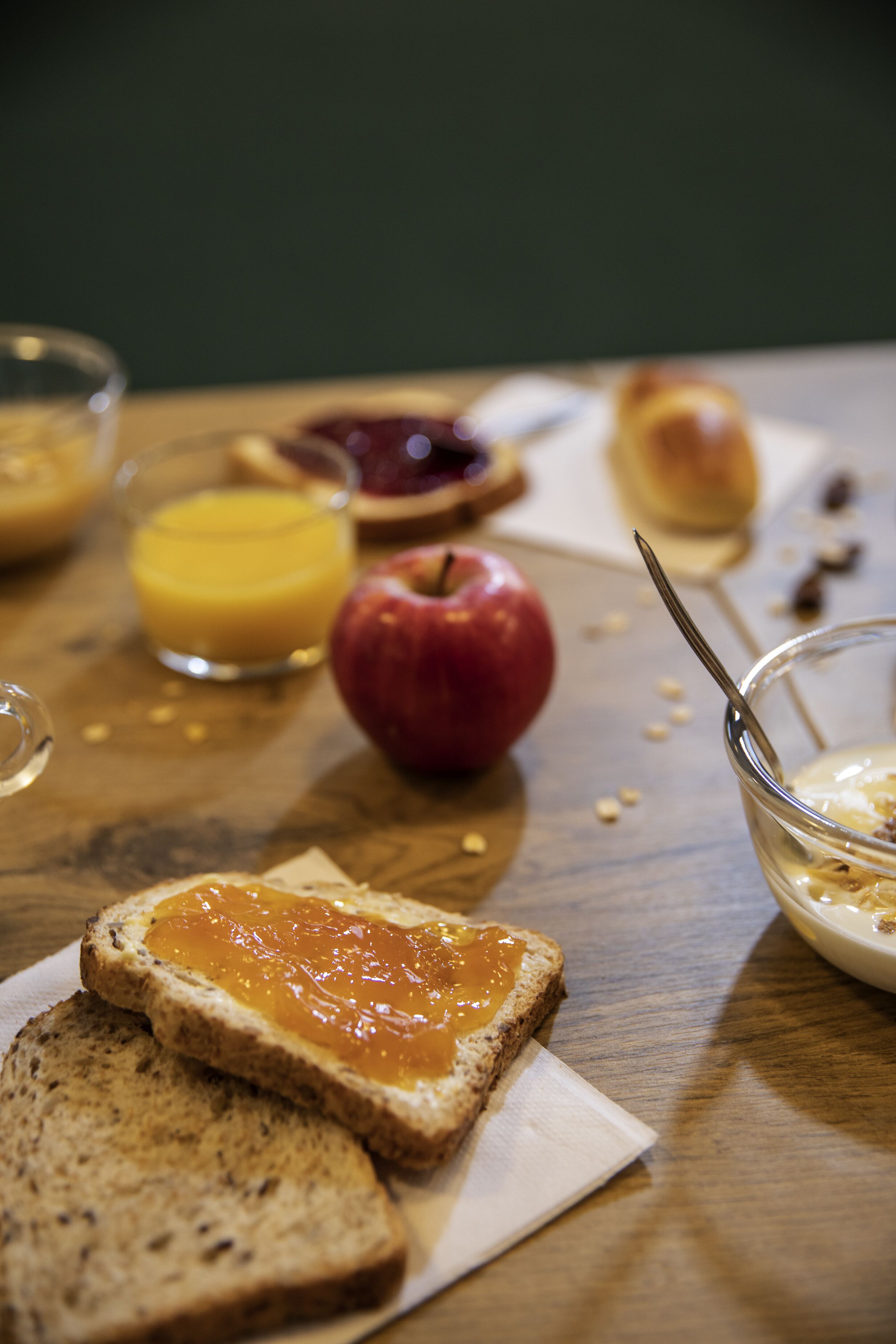 Uma mesa de madeira com itens de café da manhã, incluindo torradas com geleia, um copo de suco de laranja, uma tigela de cereal e uma maçã vermelha.