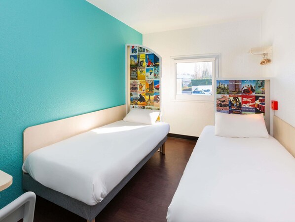 Quarto de hotel com duas camas de solteiro, janela com vista e parede decorada com cartazes de viagens.