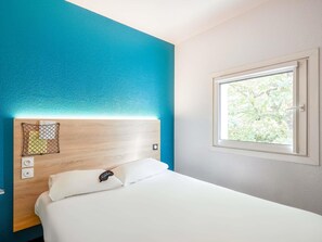 Quarto de hotel com uma cama, cabeceira com um objeto decorativo pequeno, janela com vista para vegetação e parede com detalhe azul.