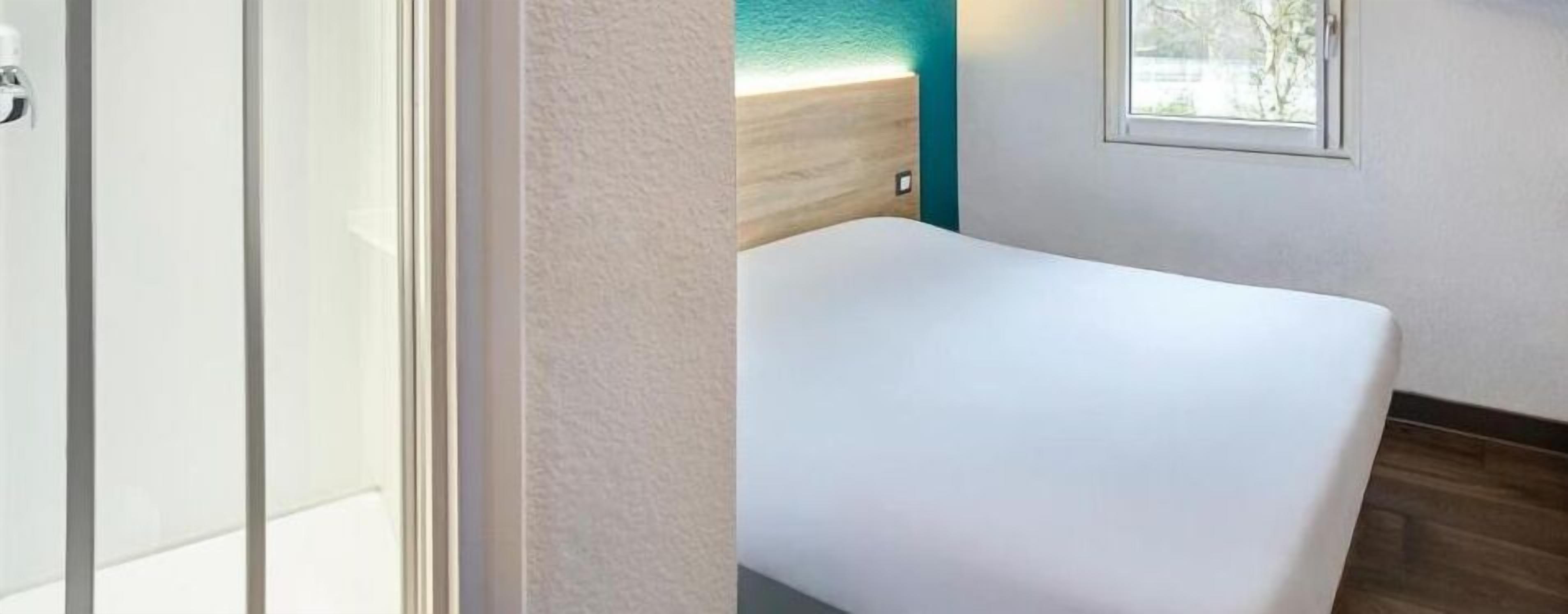 Quarto com uma cama branca, cabeceira de madeira, uma pequena janela e uma porta.