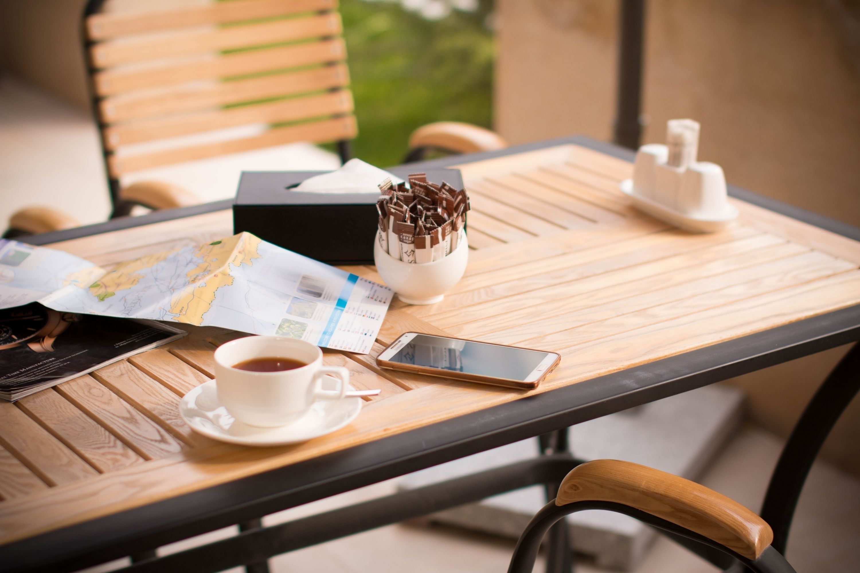 Uma mesa de madeira com um mapa, um smartphone, uma chávena de café, uma tigela de chocolates e um pequeno recipiente branco.