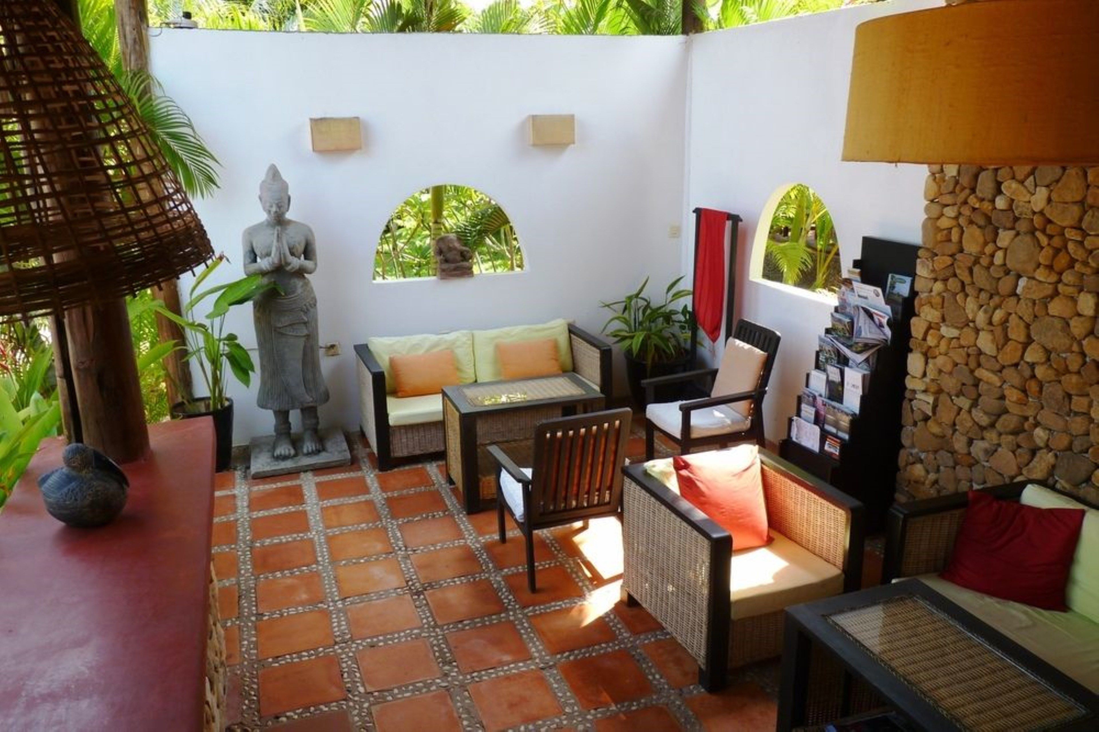 Um pátio com um sofá, cadeiras, uma mesa e uma estátua. Há janelas e uma parede de pedra.