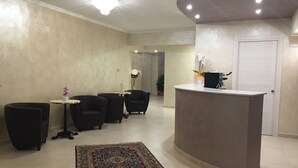 Uma área de recepção com balcão, várias poltronas, uma pequena mesa com um vaso de flores e um tapete decorativo.