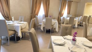 Uma sala de jantar com mesas preparadas para quatro pessoas, cada uma com toalha branca, cadeiras e talheres organizados.