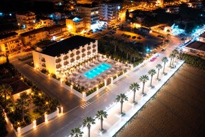 Vista aérea de um hotel com piscina, rodeado por palmeiras e uma rua bem iluminada.