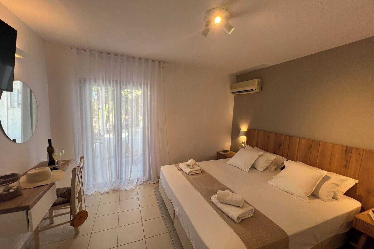 Quarto de hotel com uma cama grande, uma escrivaninha, uma cadeira, um espelho e uma janela com cortinas.