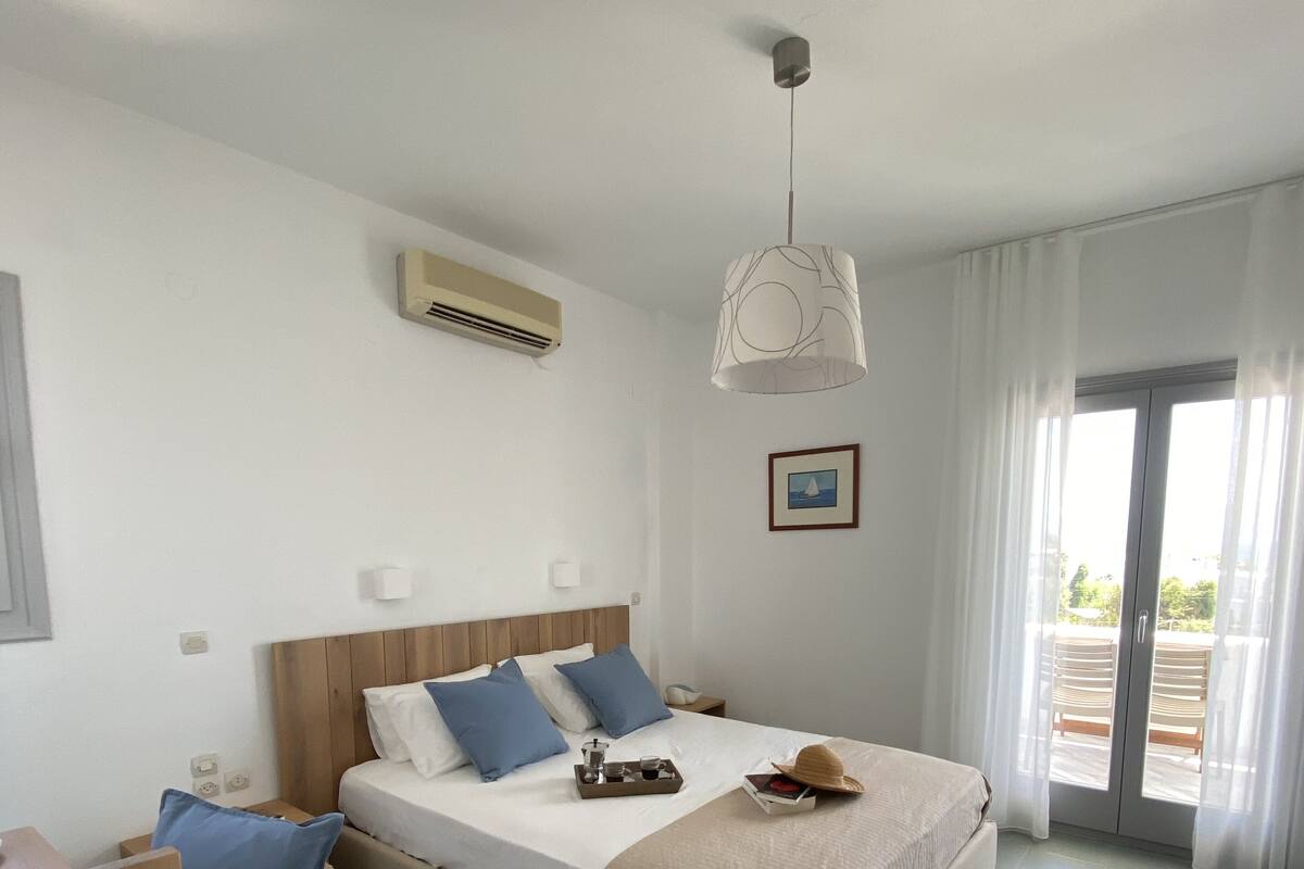 Quarto de hotel com cama, escrivaninha, cadeira, janela com cortinas, ar condicionado e um quadro na parede.