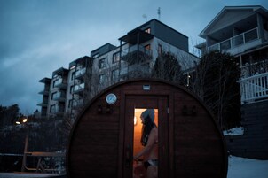 Uma estrutura de sauna de madeira com porta, rodeada por edifícios residenciais.