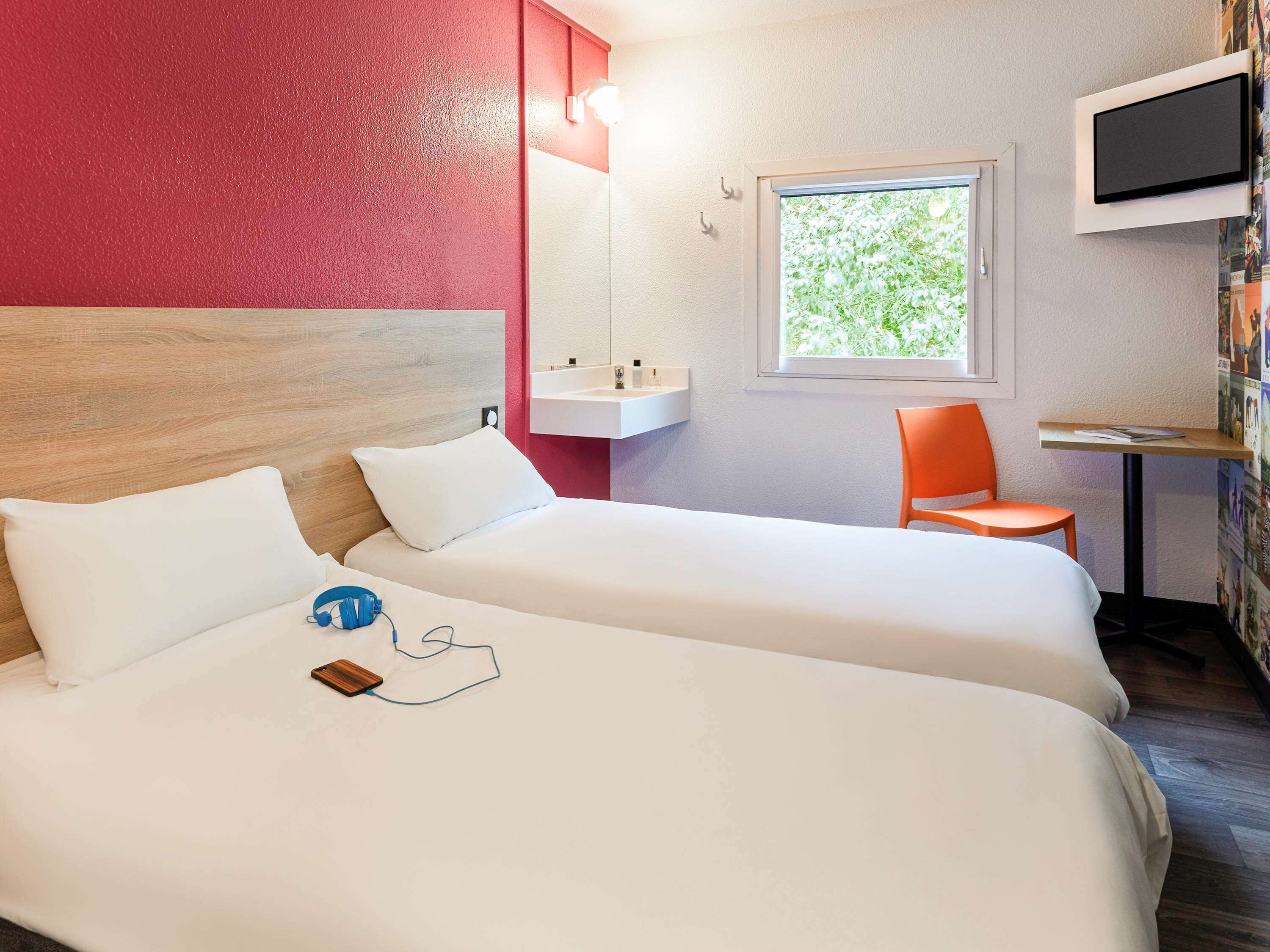 Quarto de hotel com duas camas, uma escrivaninha, uma cadeira, uma TV e uma janela com vista para o verde.