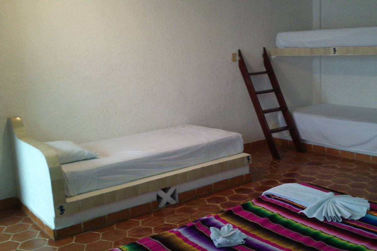Quarto pequeno com uma cama de solteiro, uma escada de madeira encostada na parede, um tapete listrado e colorido no chão e uma cama branca com um único travesseiro.