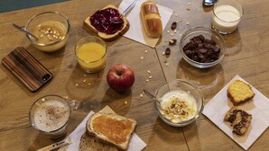 Uma mesa de madeira com itens de café da manhã, incluindo torradas, frutas, cereais e um copo de suco de laranja.