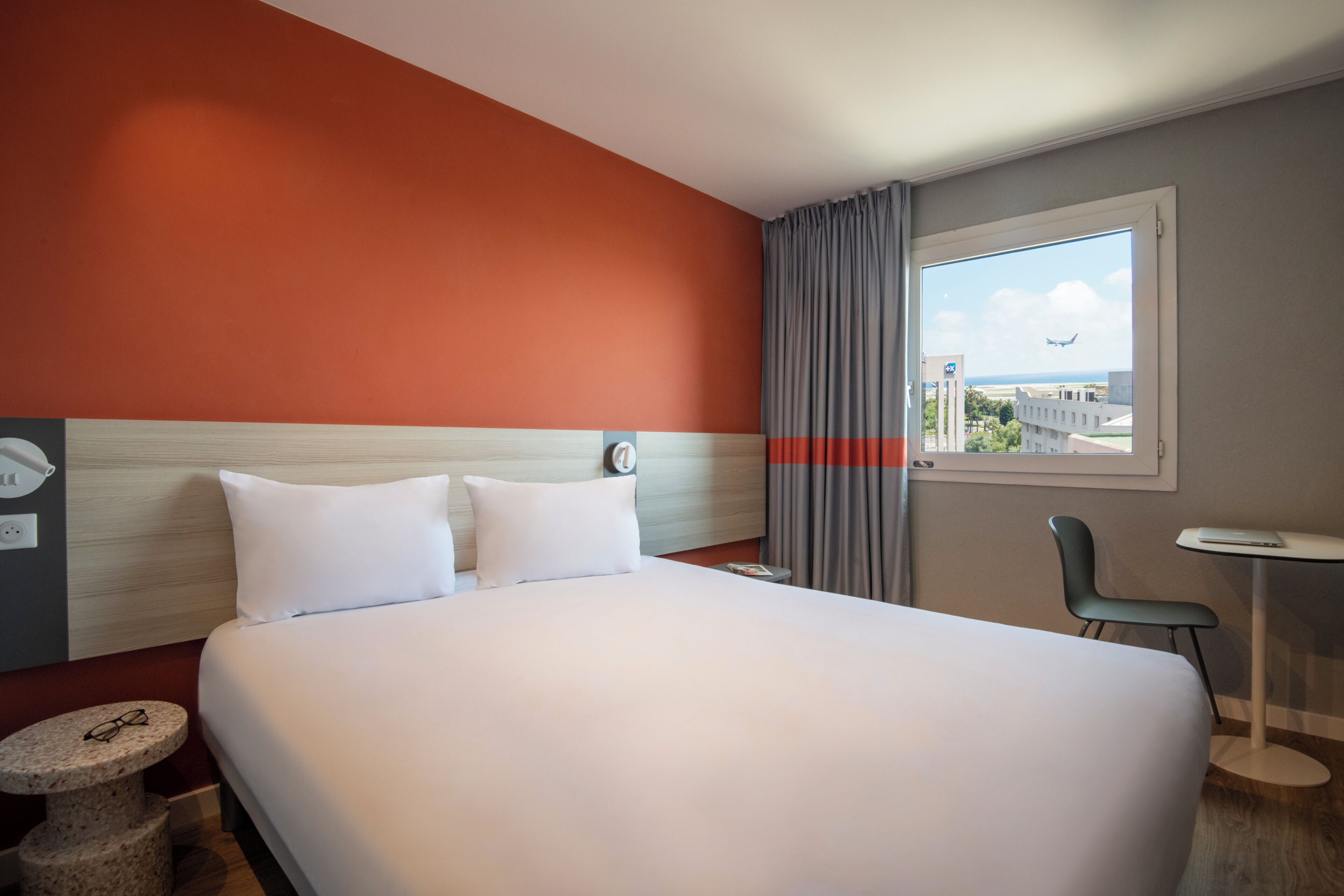 Quarto de hotel com uma cama grande, uma mesinha redonda, uma cadeira, uma janela com vista para prédios e um avião, e um relógio na parede.