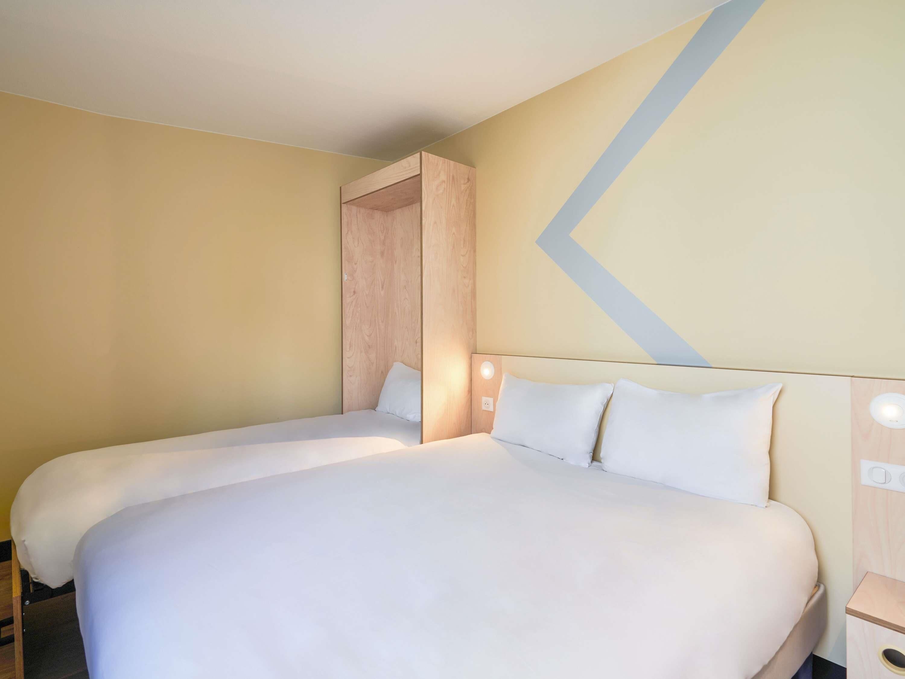 Quarto de hotel com duas camas, cabeceira de madeira, parede em tom claro e um design geométrico.