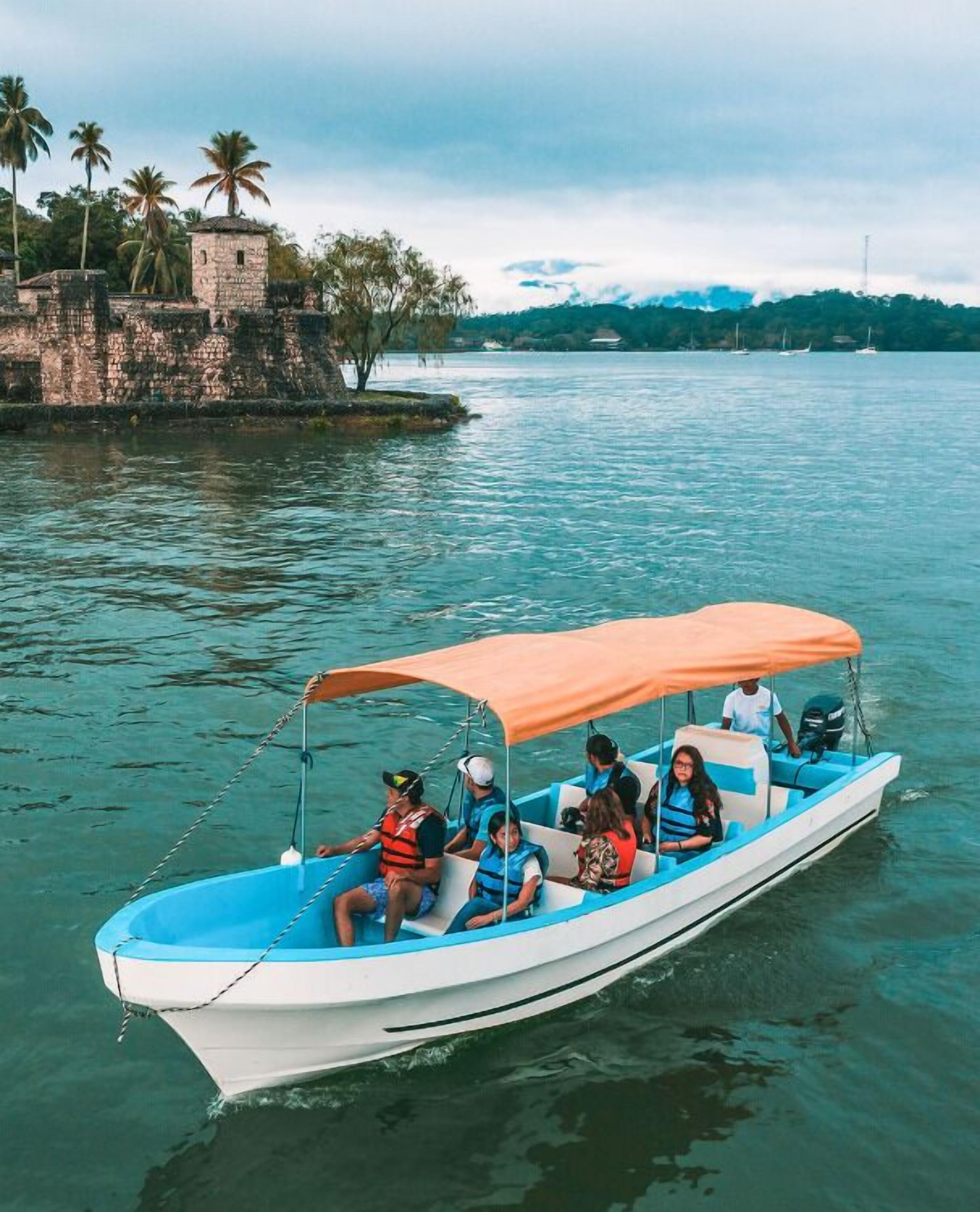 Um barco com passageiros, uma cobertura e uma estrutura de pedra ao fundo.