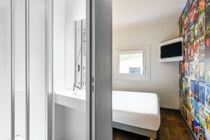 Quarto com uma cama, janela, televisão presa na parede e parede coberta por cartazes coloridos.