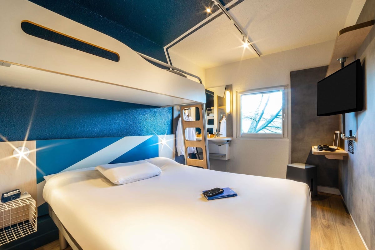 Quarto de hotel moderno com uma cama grande, banheiro pequeno, televisão fixada na parede e janela com luz natural.