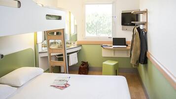 Quarto de hotel compacto com beliches, uma escrivaninha com um laptop, um banquinho pequeno e uma janela com vista para árvores.