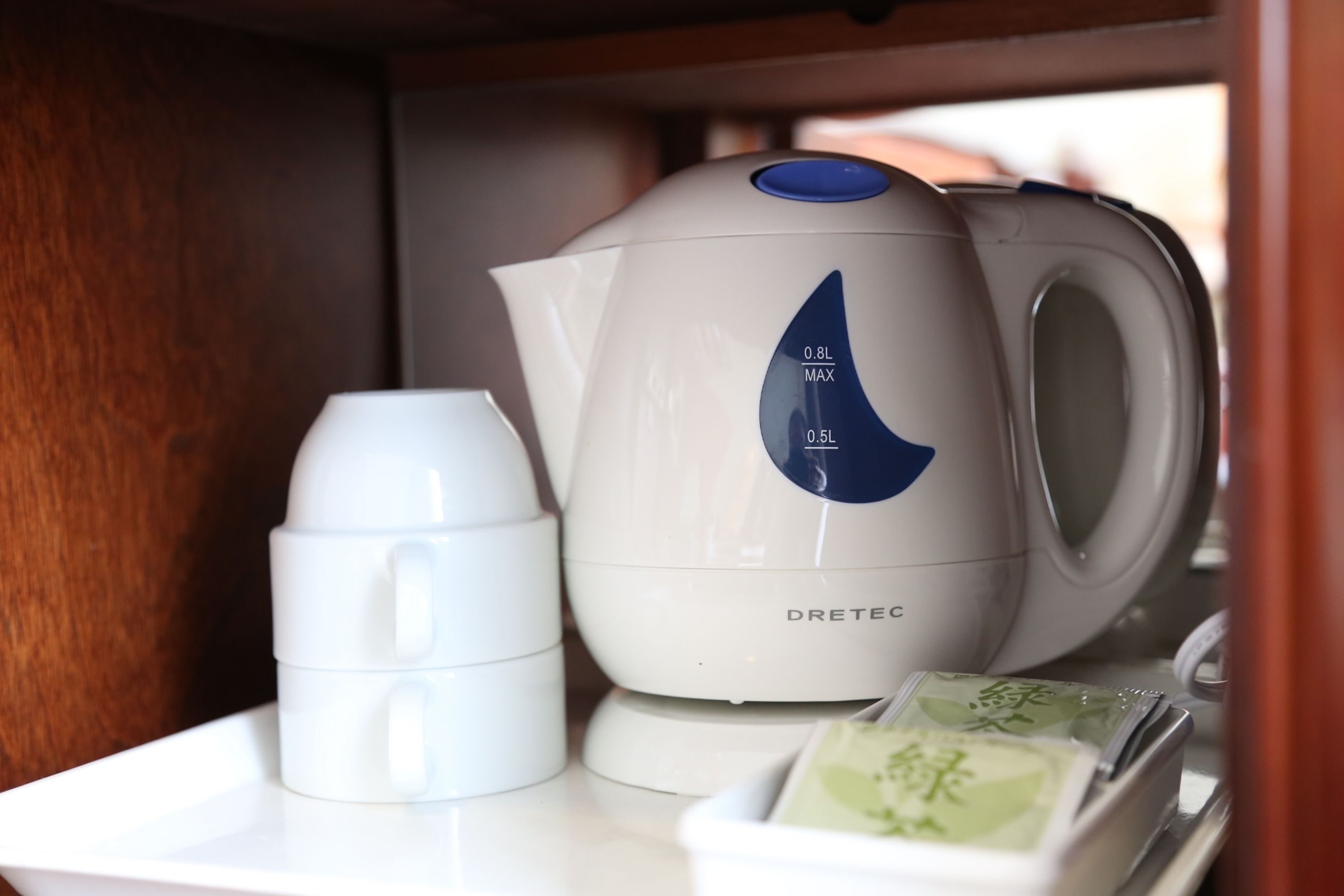 Uma chaleira branca com etiqueta azul, uma caneca branca e um pequeno recipiente com chá verde sobre uma bandeja branca.