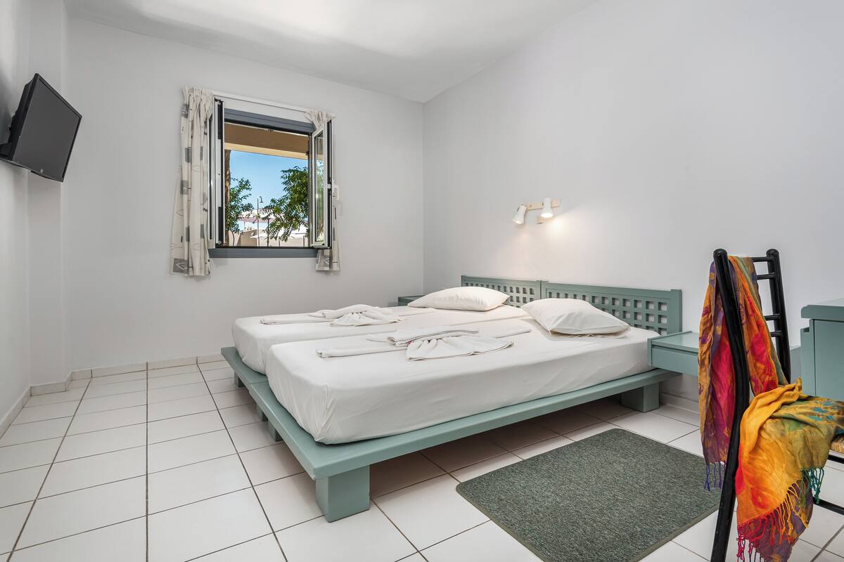 Quarto com cama, televisão fixada na parede, janela com cortinas e mesa de cabeceira com abajur.