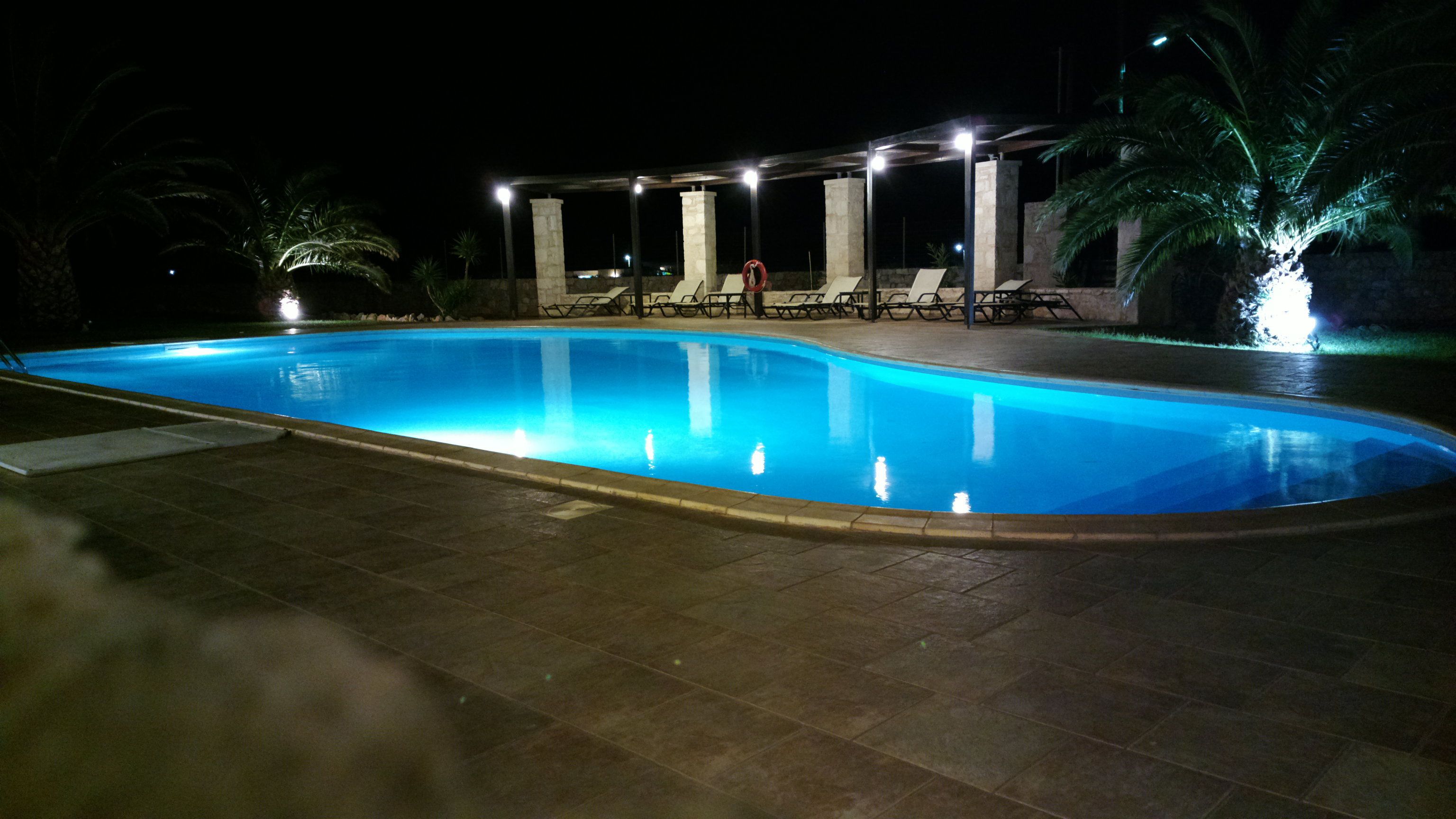 Piscina à noite com deck ladrilhado, cadeiras de descanso e palmeiras.