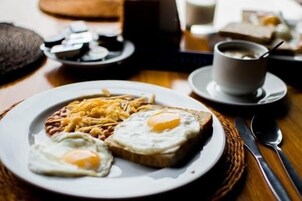 Prato de café da manhã com ovos e queijo no pão, uma xícara de café e um pratinho com doces sobre uma mesa.