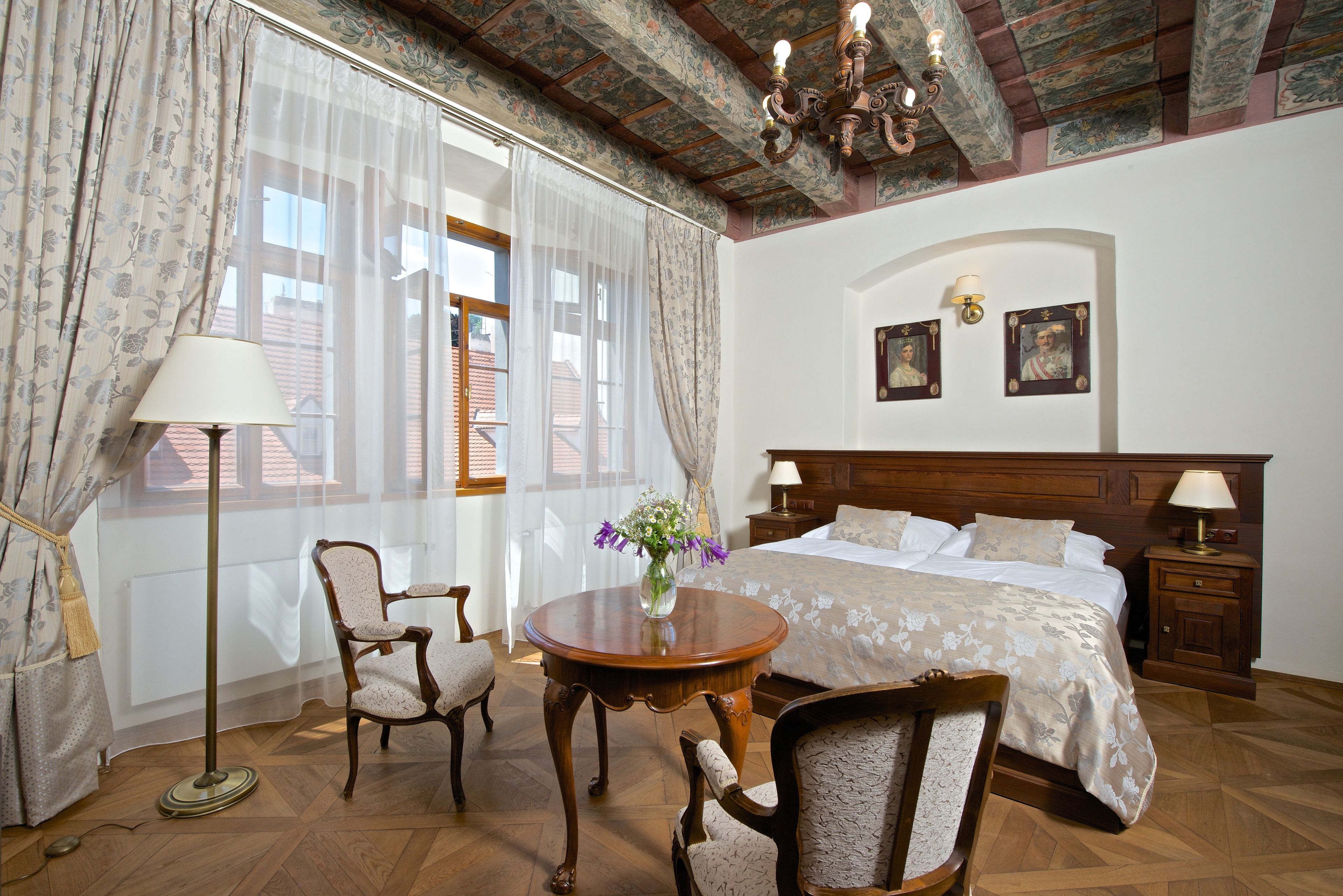 Quarto de hotel com uma cama grande, duas mesas de cabeceira, uma mesa redonda de madeira com cadeiras, um abajur e uma janela com cortinas finas.