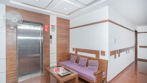 Um hall com um sofá de madeira, uma mesa de vidro com revistas, um ventilador e um elevador.